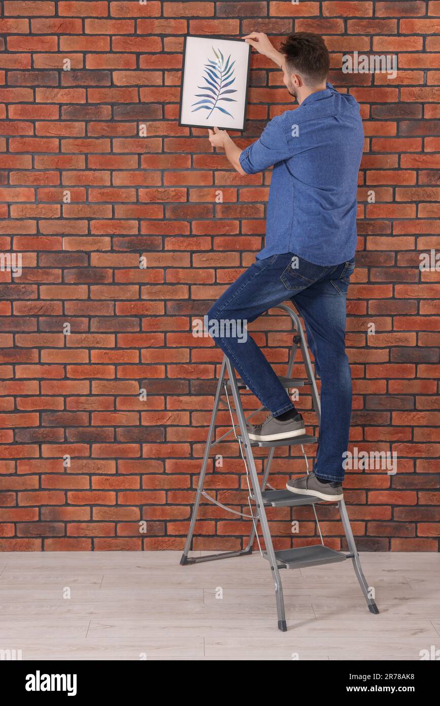 Man on stepladder hanging picture near red brick wall Stock Photo - Alamy