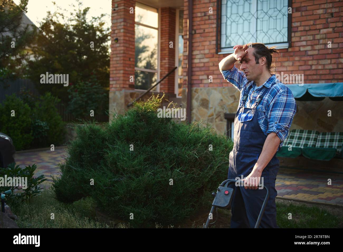 Male gardener in blue working uniform wiping sweat from forehead, tired ...