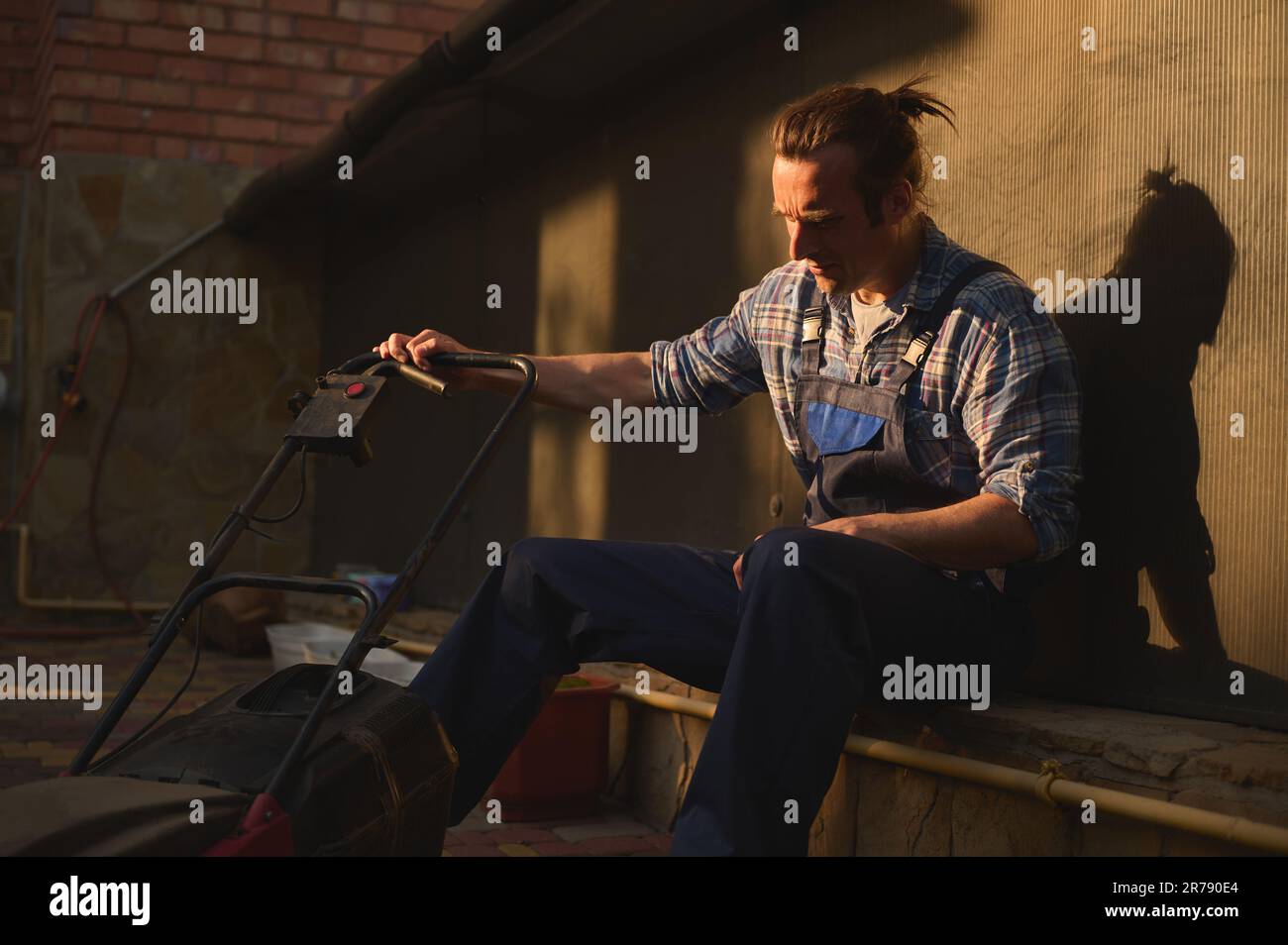 Portrait of an overworked gardener in blue overalls with a lawn mower ...