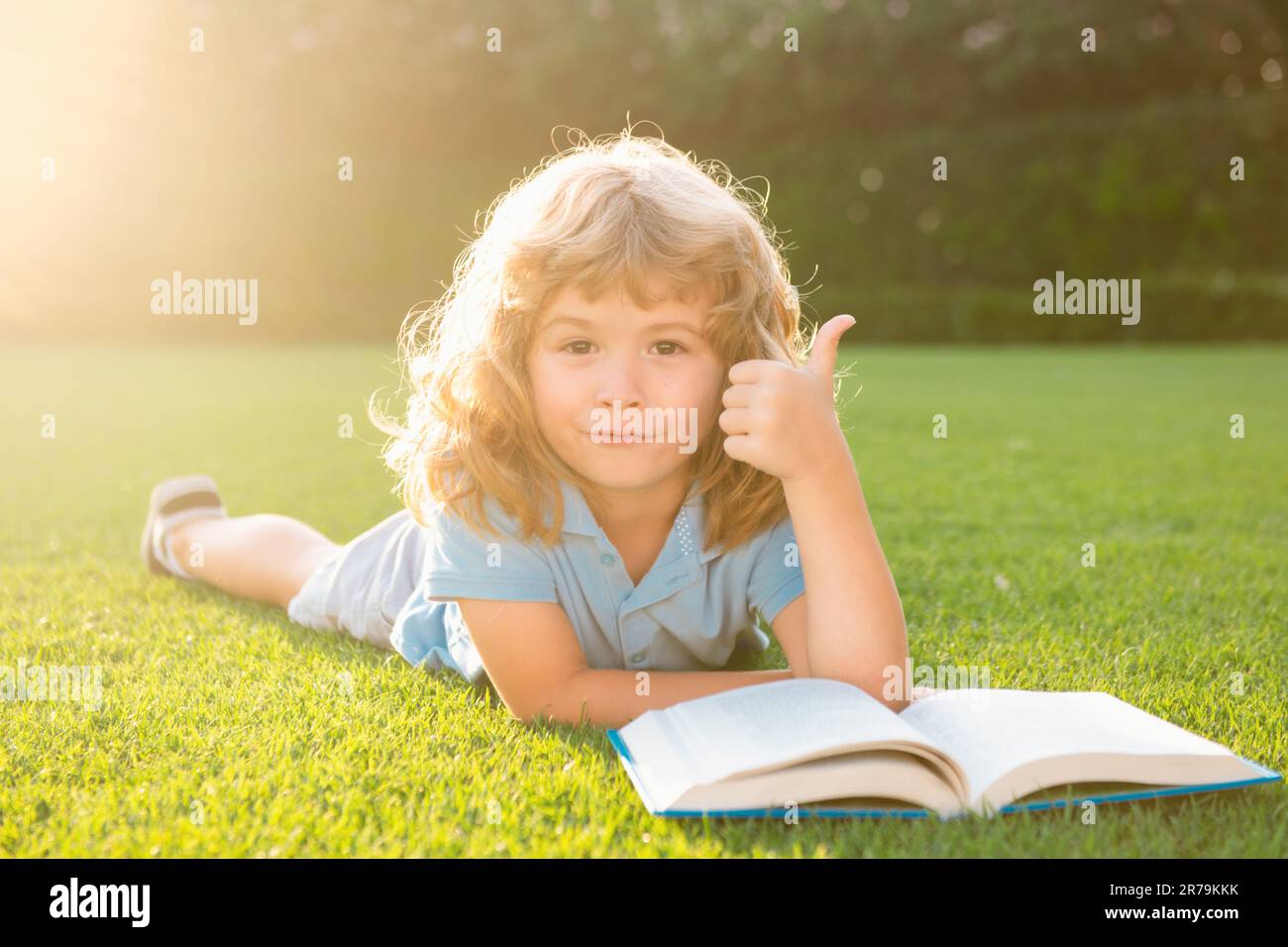 Cute boy with thumbs up reading book on green grass. Summer outdoor ...