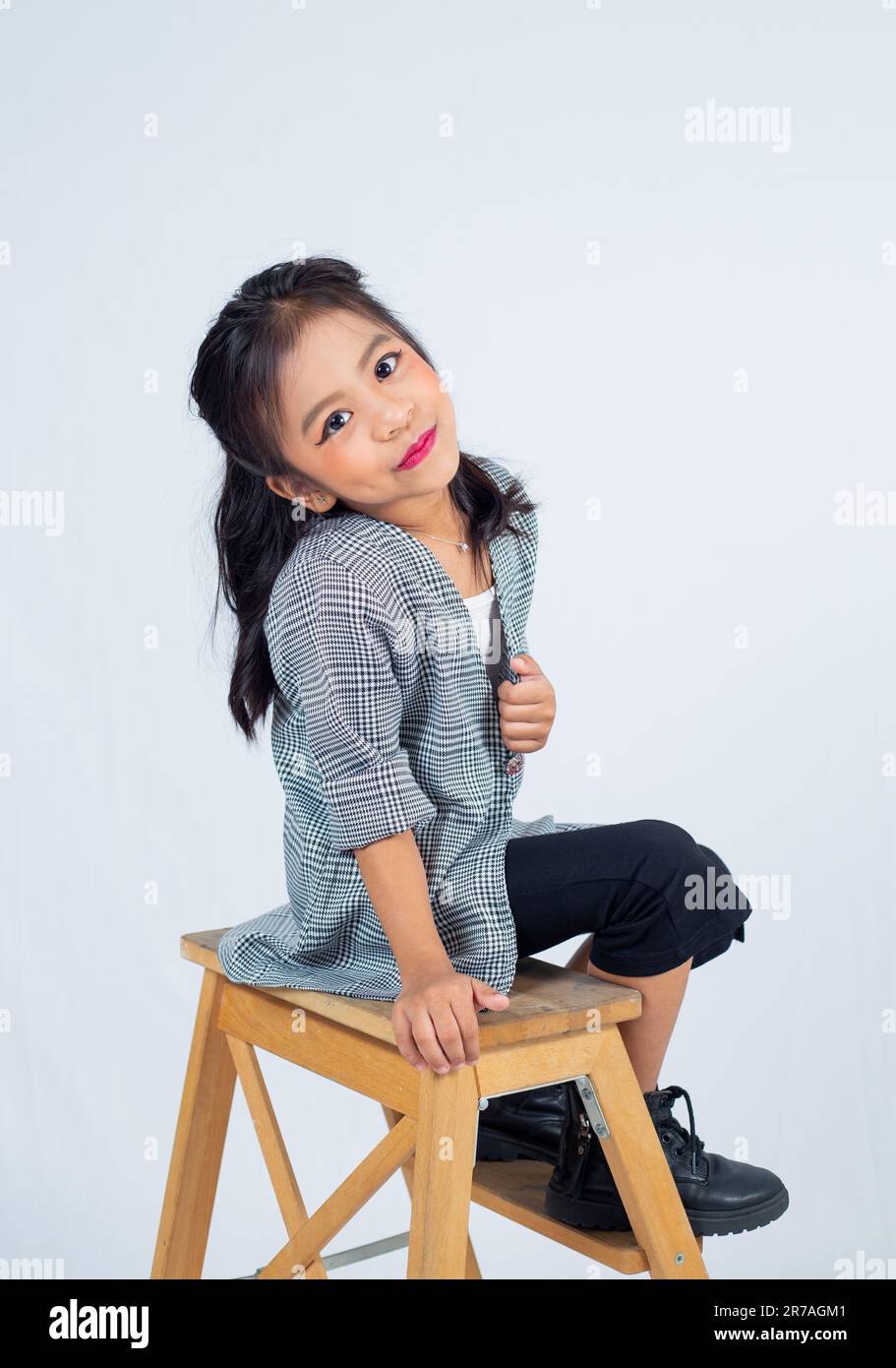 A young female child is perched atop a white stool against a plain ...