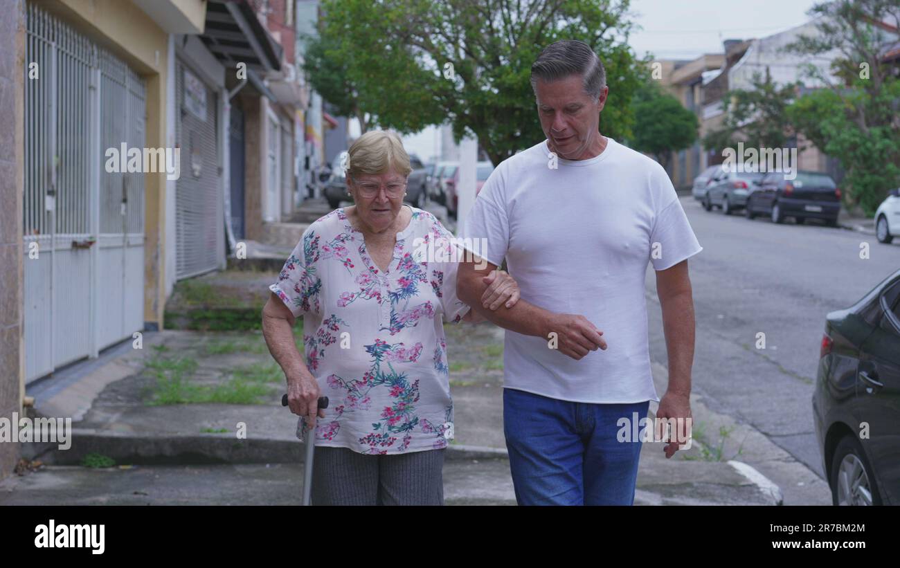 Adult son assisting elderly mother to walk in sidewalk. Senior woman ...