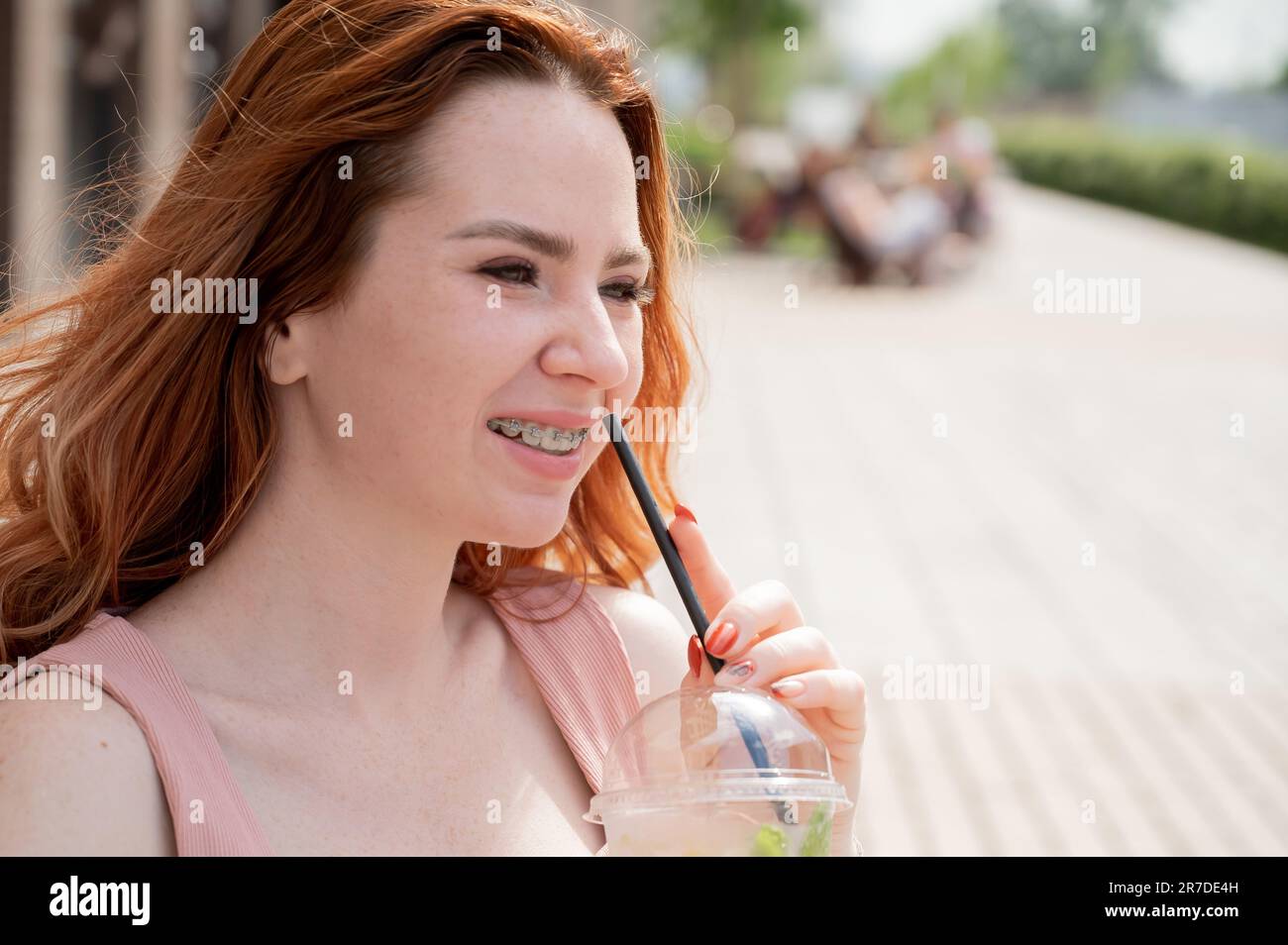 Young beautiful red-haired woman with braces drinks cooling lemonade ...