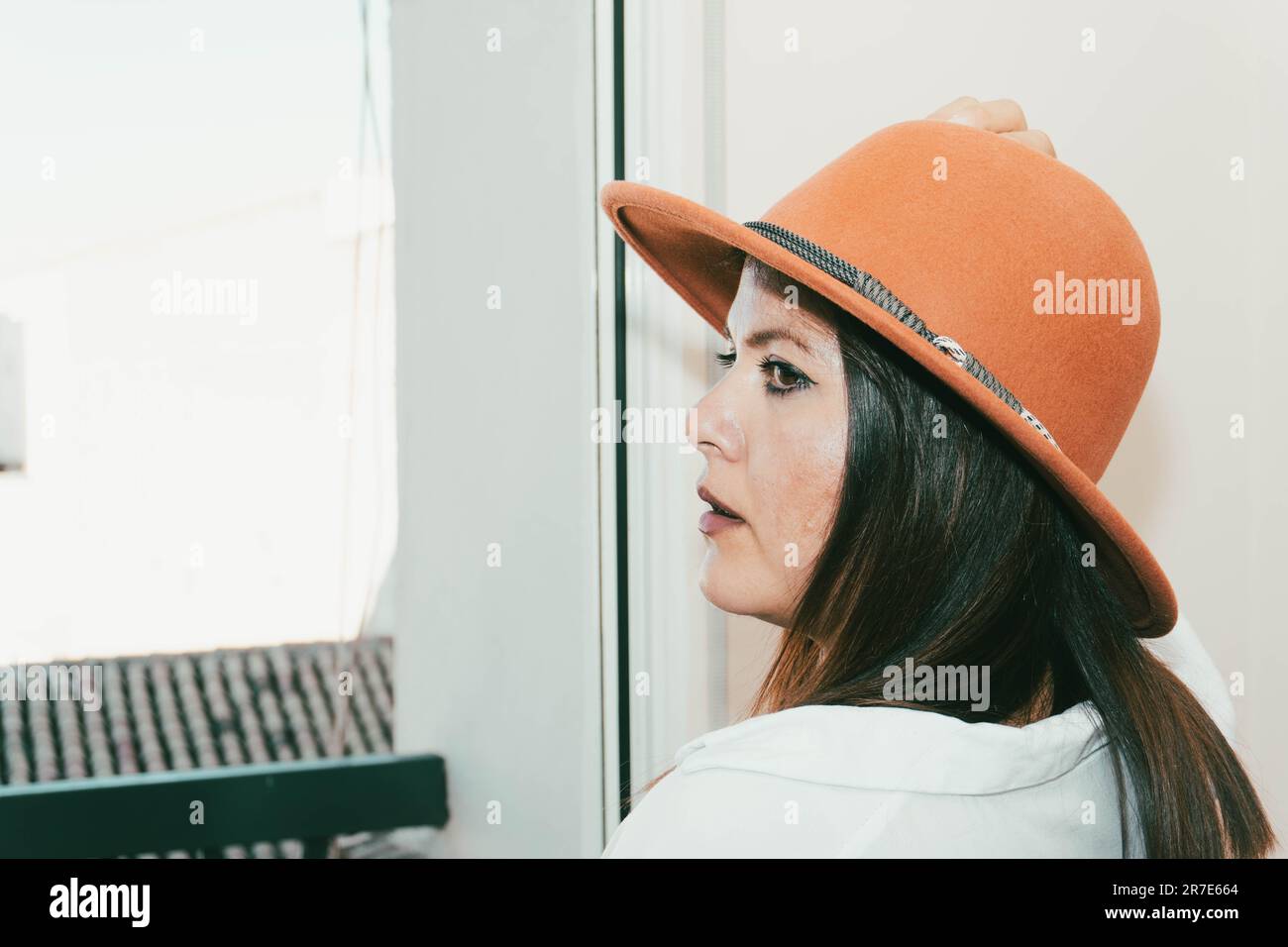 A mature woman with an orange hat stands at a window looking backwards ...