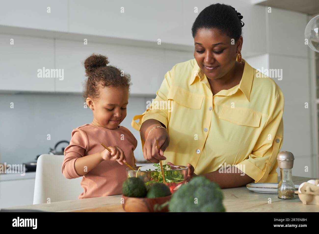 African American mom cooking vegetable salad in the kitchen with little ...