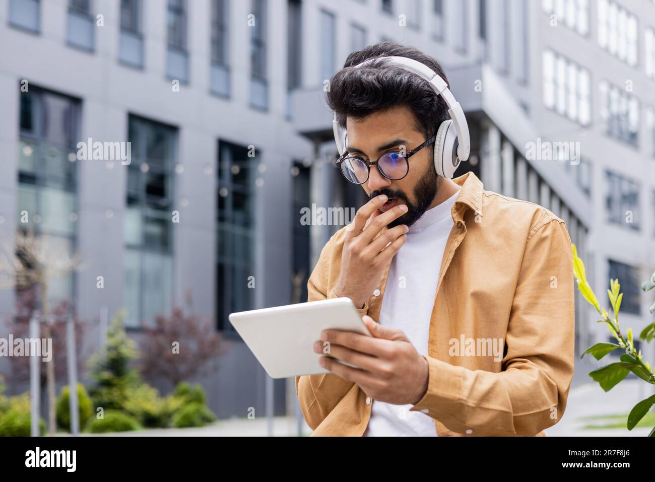 Young man with headphones and tablet computer sitting outside office building on park bench ...