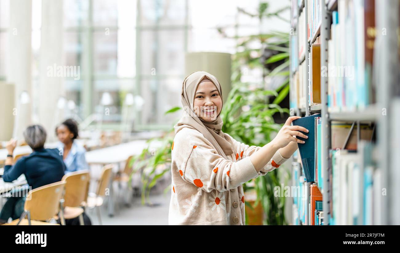 Asian muslim female student picking book from bookshelf in library ...