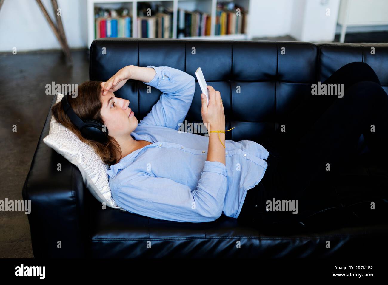 Young woman stretched out on leather sofa with headphones, using mobile ...