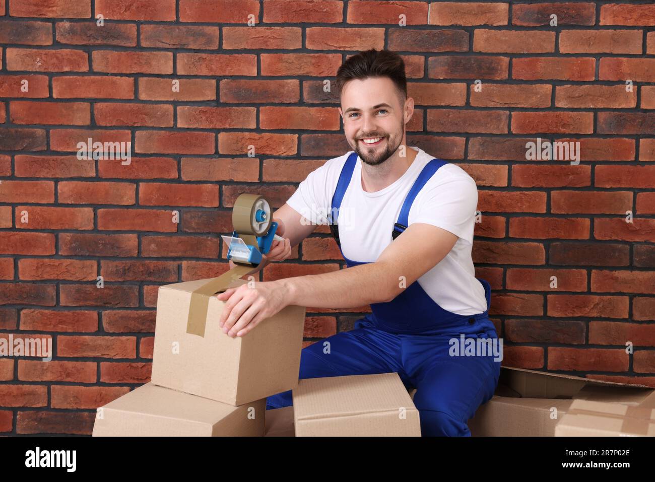 Smiling worker taping box with adhesive tape dispenser near brick wall ...