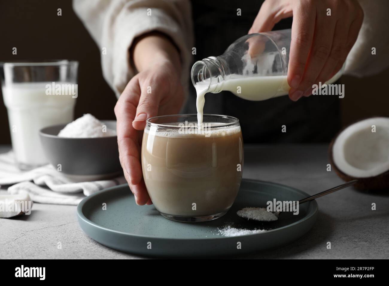 Woman pouring coconut milk into glass of coffee at light grey table ...