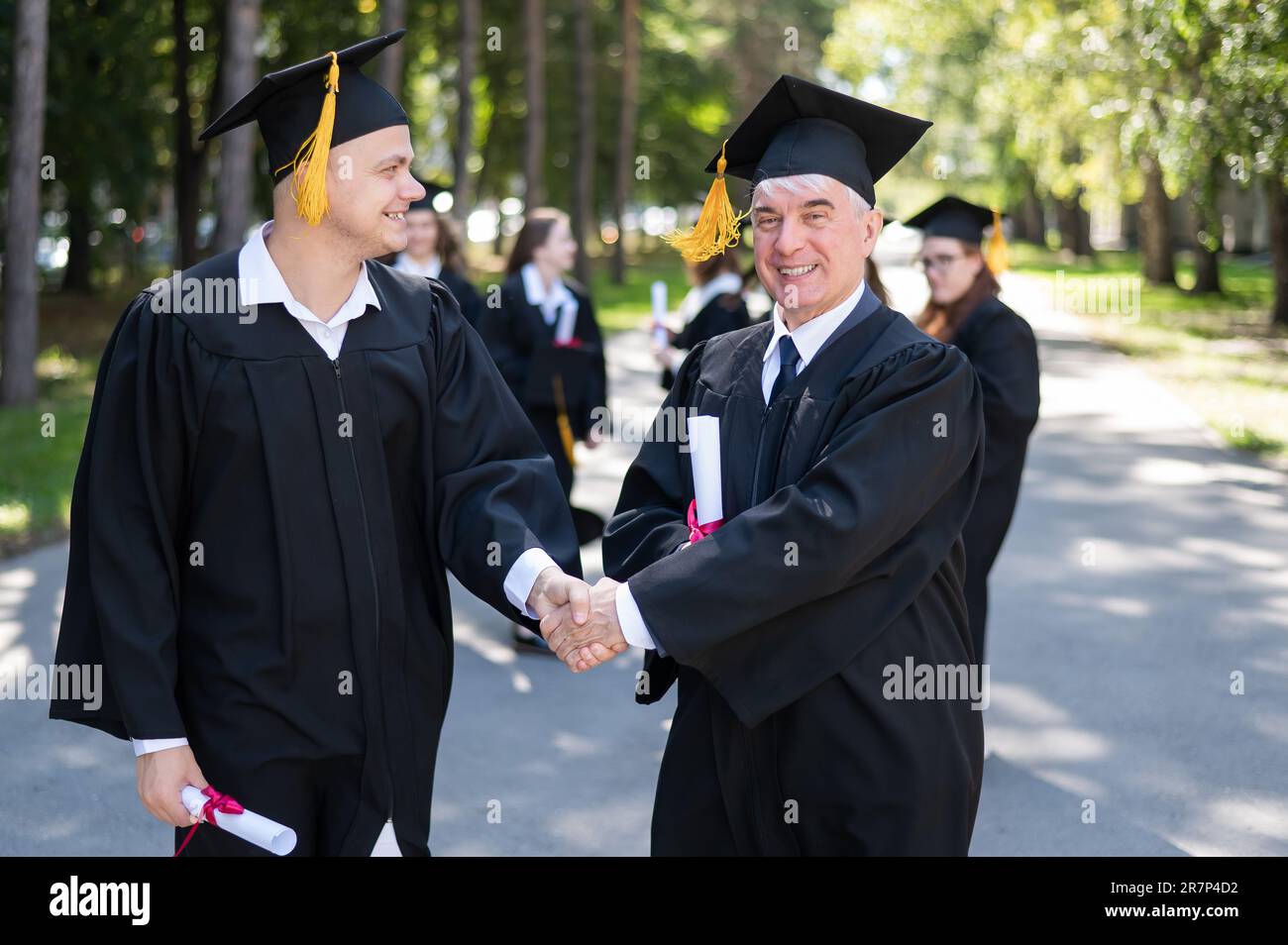 A group of graduates in robes outdoors. An elderly man and a young guy ...