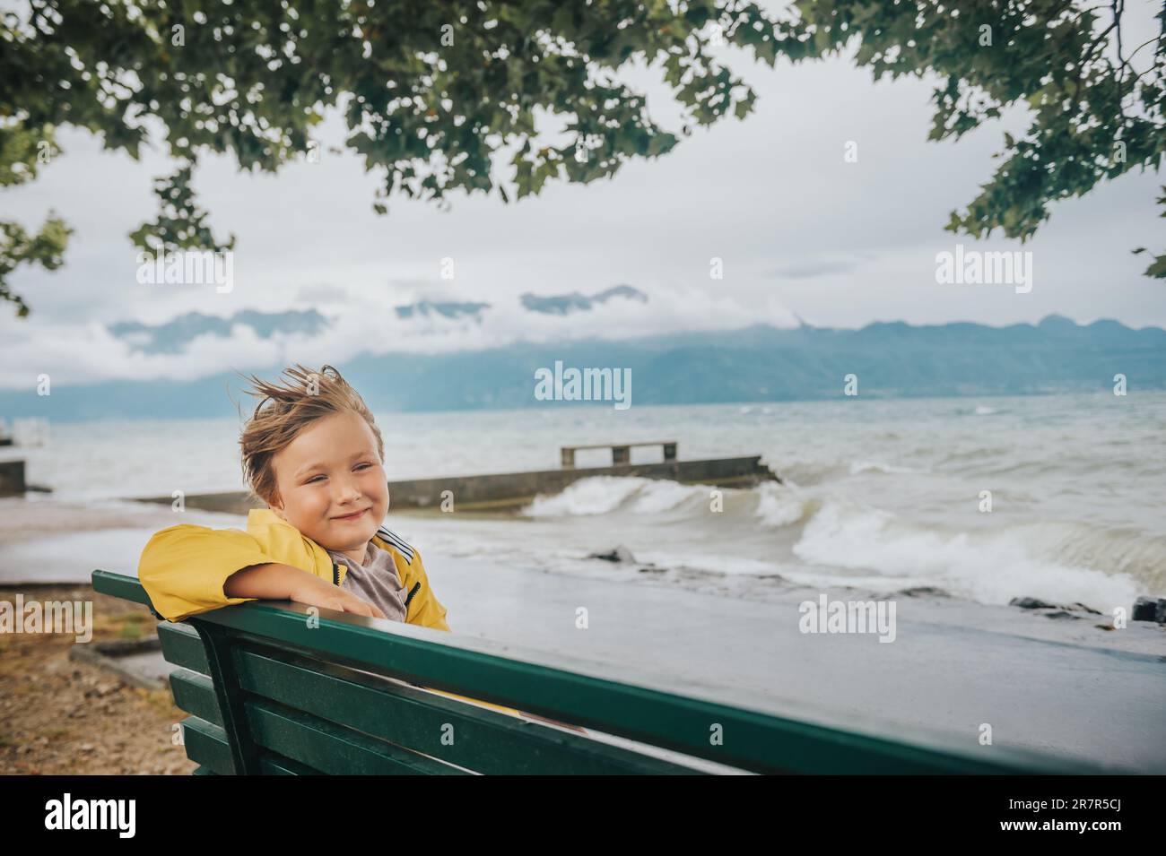 Cute little boy resting on a bench by the lake on a very windy day ...