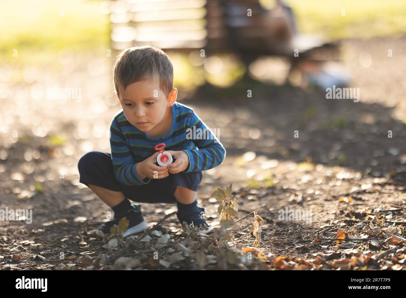 Little boy crouched in the park holding soap bubbles stick. Mid shot ...