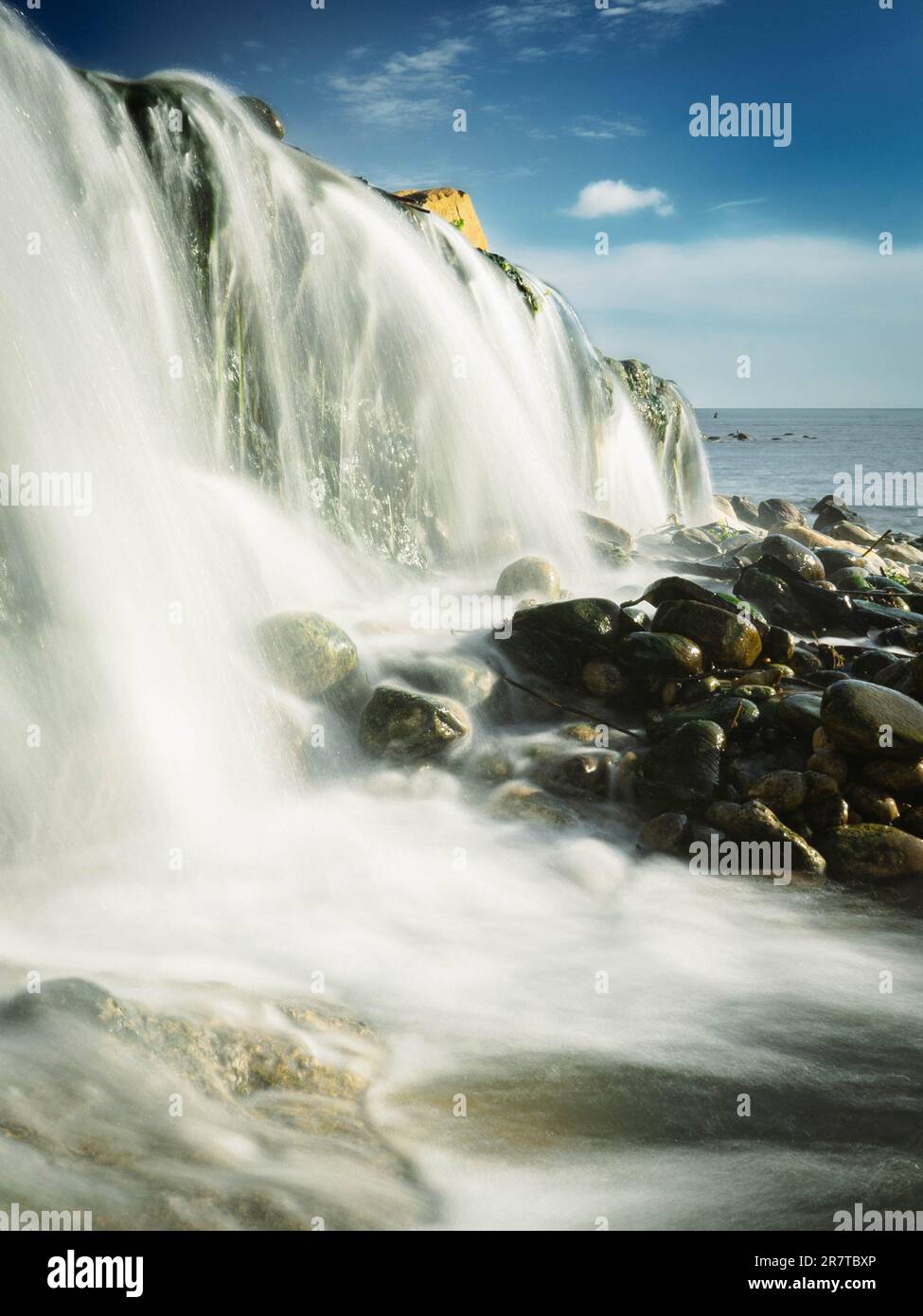 A majestic waterfall cascades down a steep rock face over the stony sea ...