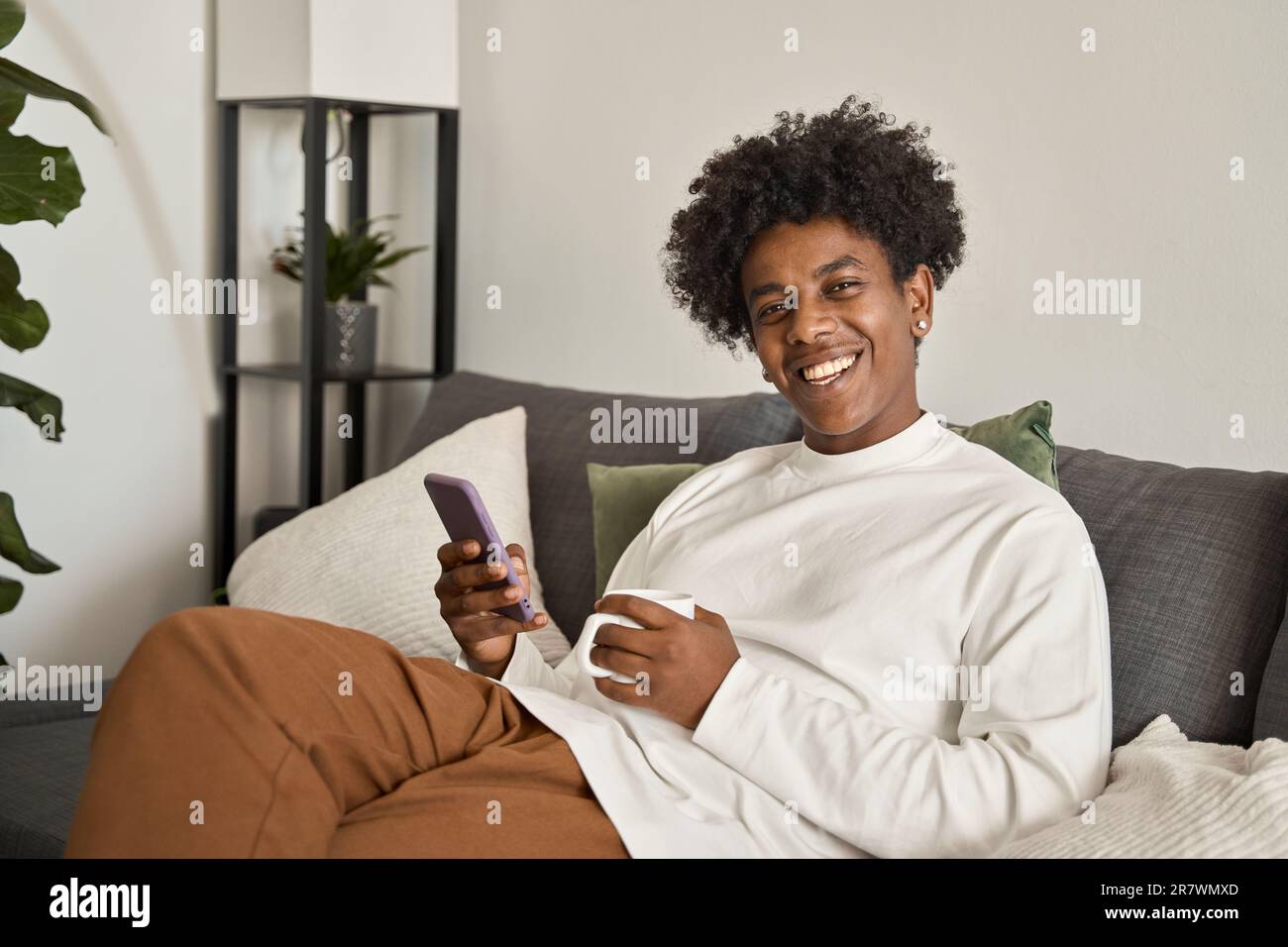 Smiling gen z African American teen sitting on couch using smartphone ...