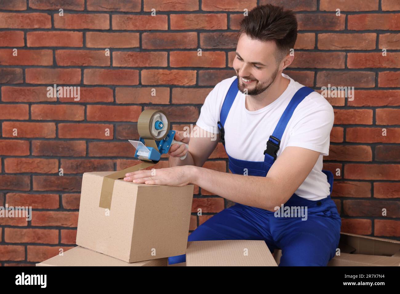 Smiling worker taping box with adhesive tape dispenser near brick wall ...