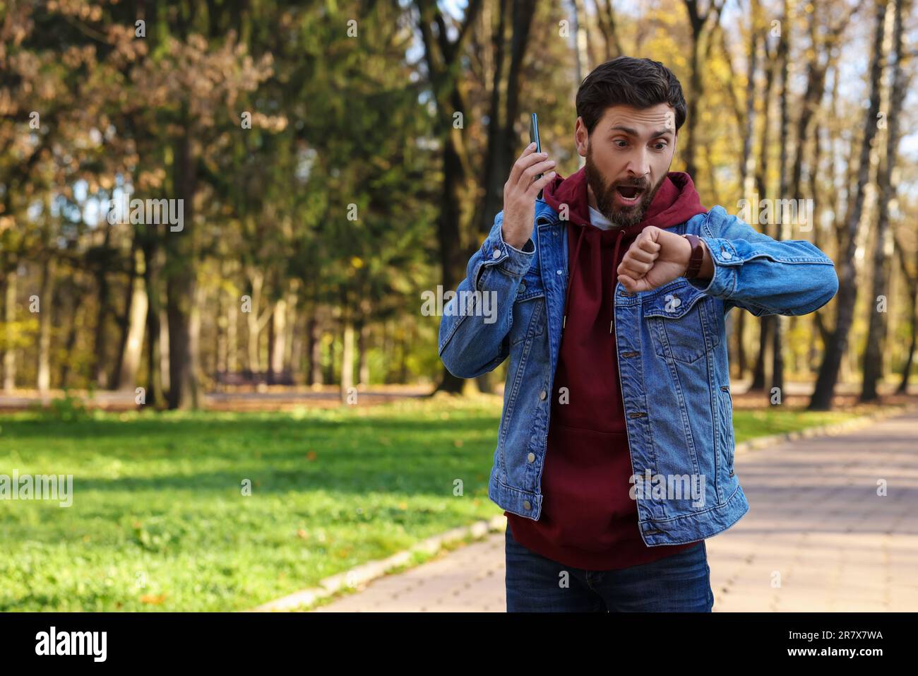 Emotional man talking by smartphone and checking time on watch in park ...