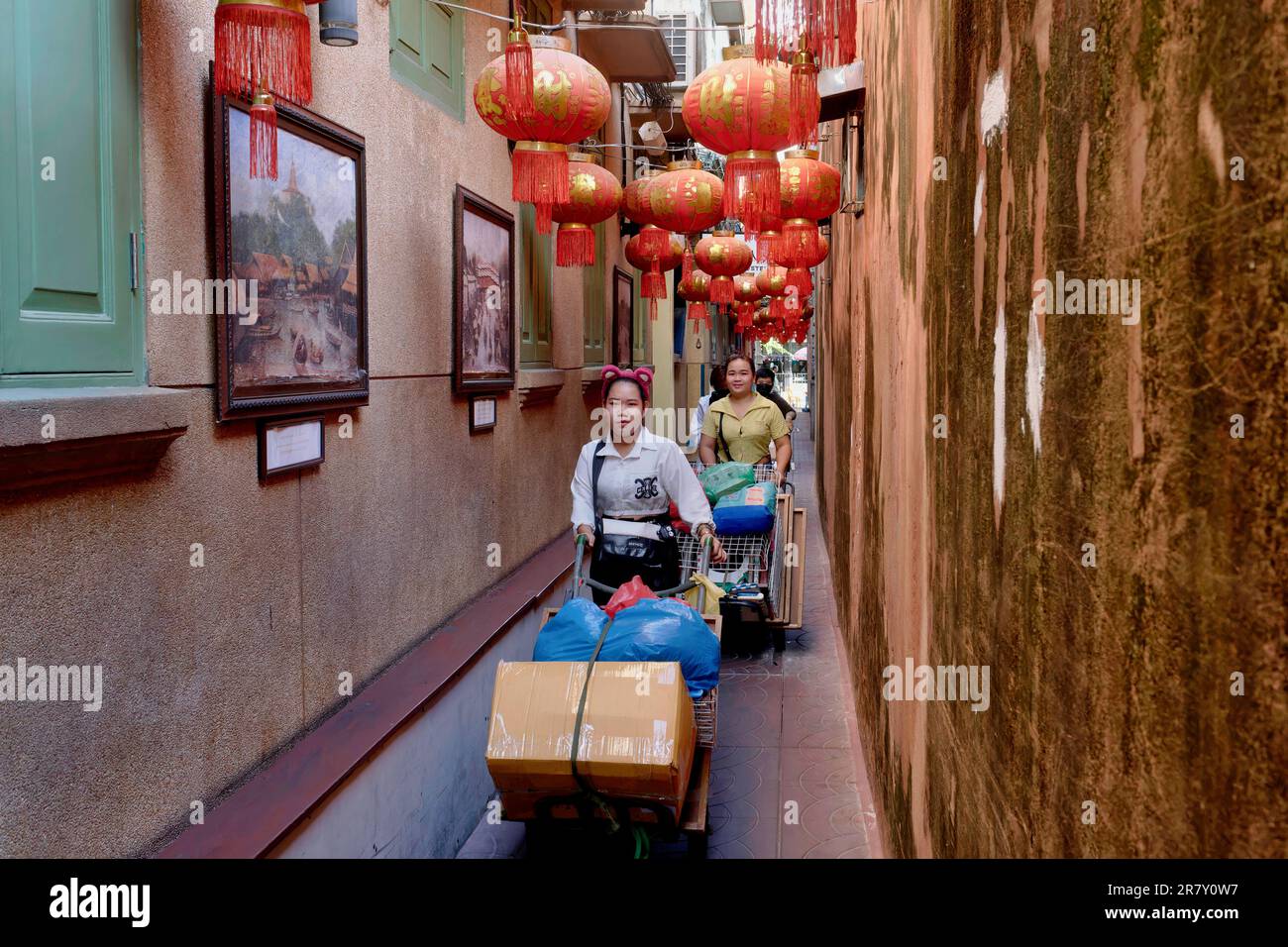 Two women are pushing trolleys laden with goods through a narrow lane ...
