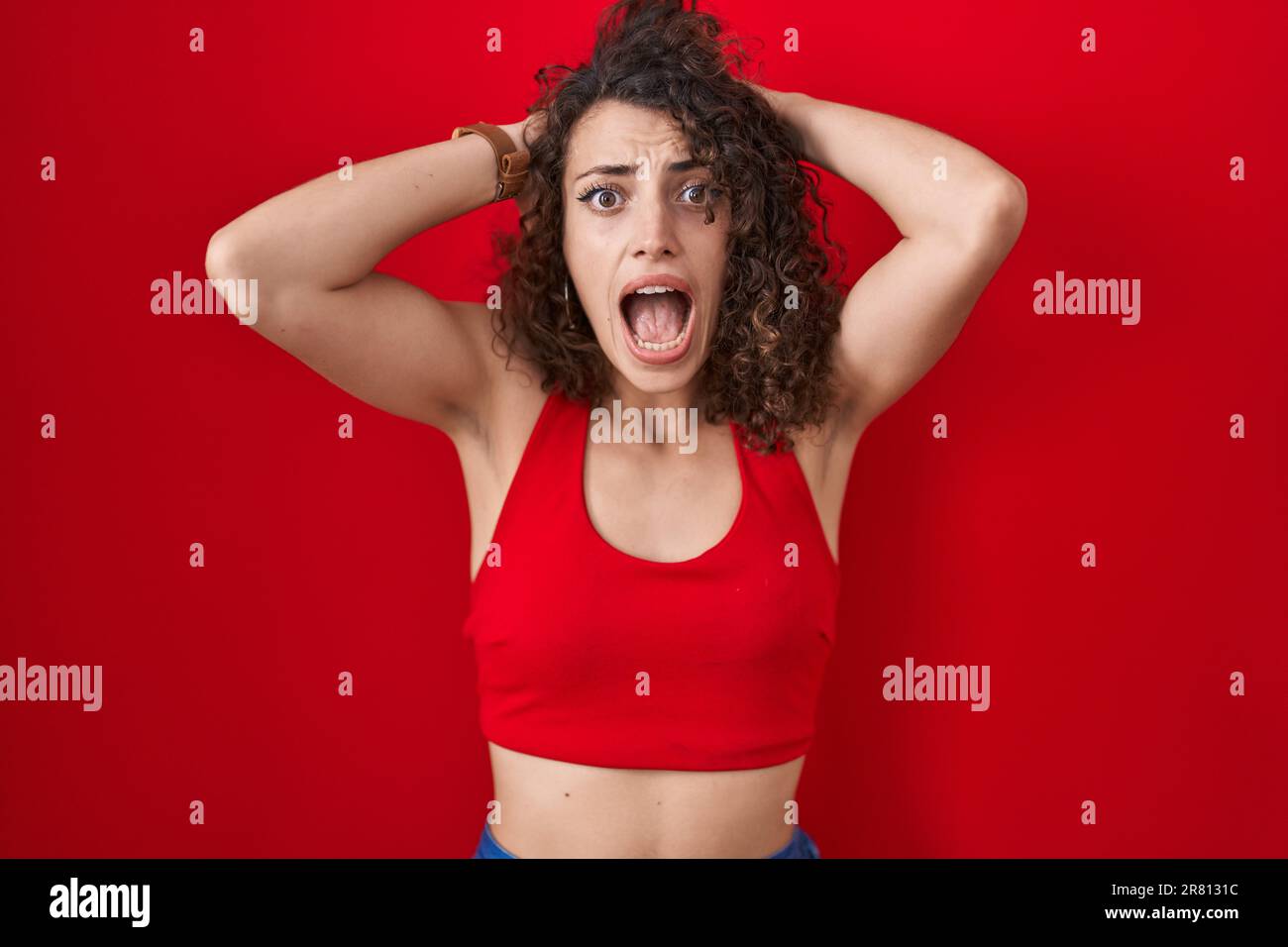 Hispanic woman with curly hair standing over red background crazy and ...