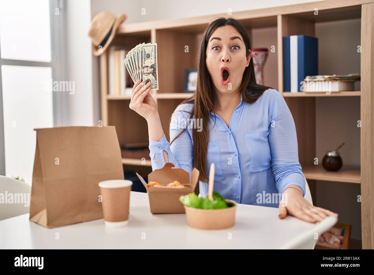 Young brunette woman eating take away food at home holding money scared ...