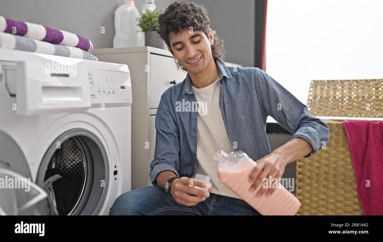 Young hispanic man washing clothes pouring detergent smiling at laundry ...