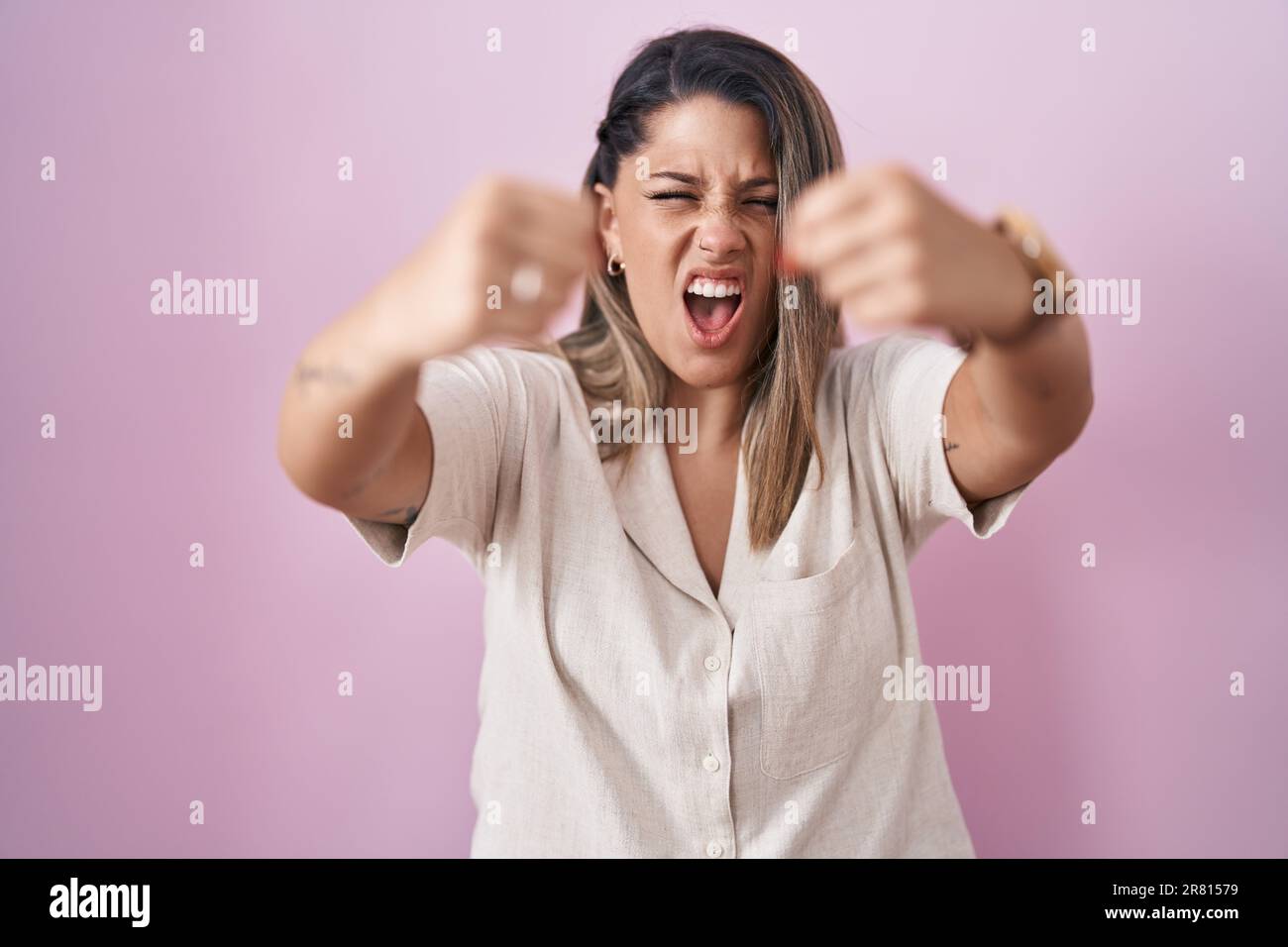 Blonde woman standing over pink background angry and mad raising fists ...