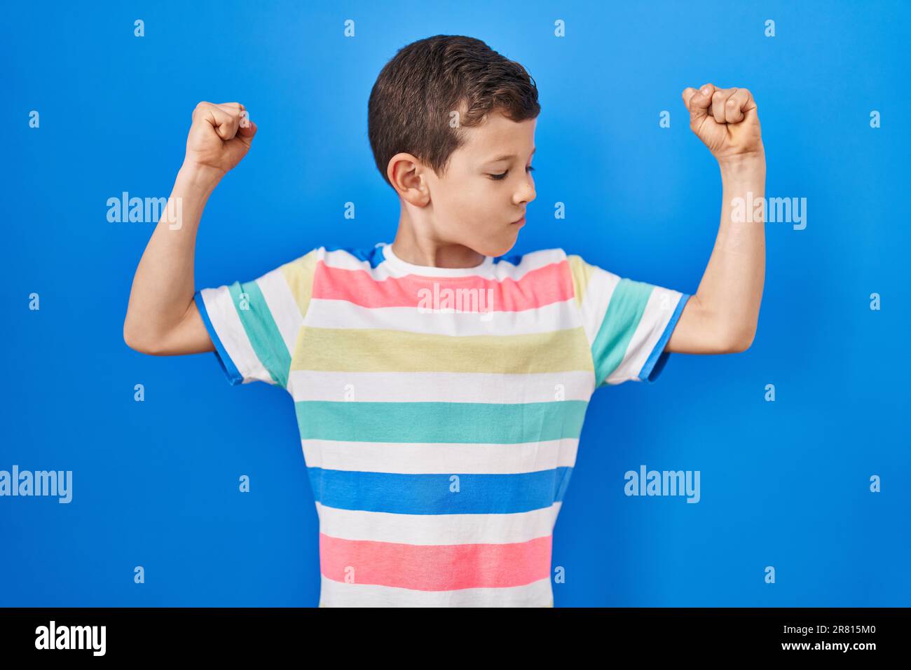 Young caucasian kid standing over blue background showing arms muscles ...
