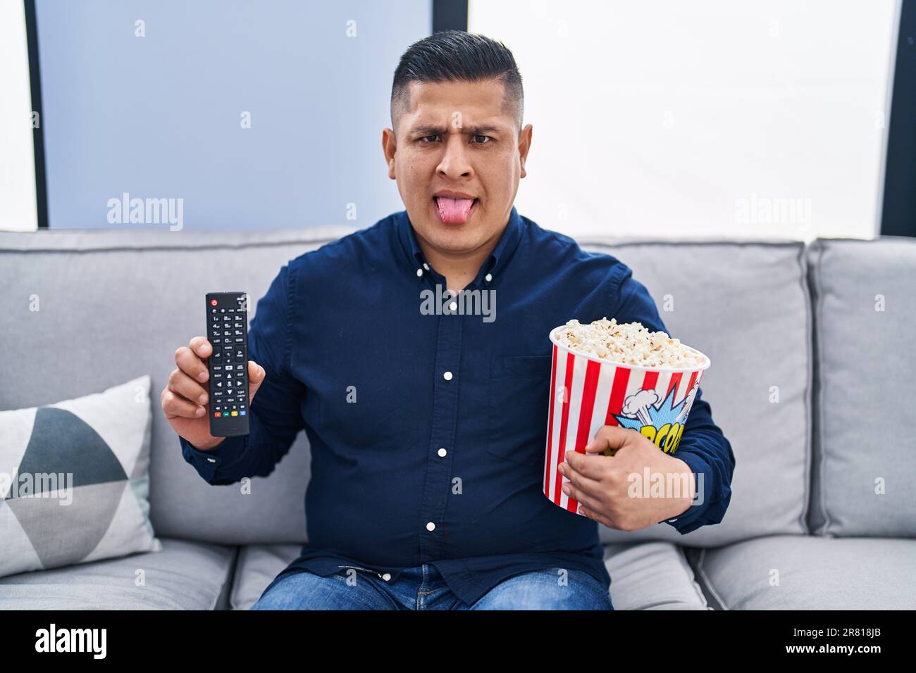 Hispanic young man eating popcorn using tv control sticking tongue out ...