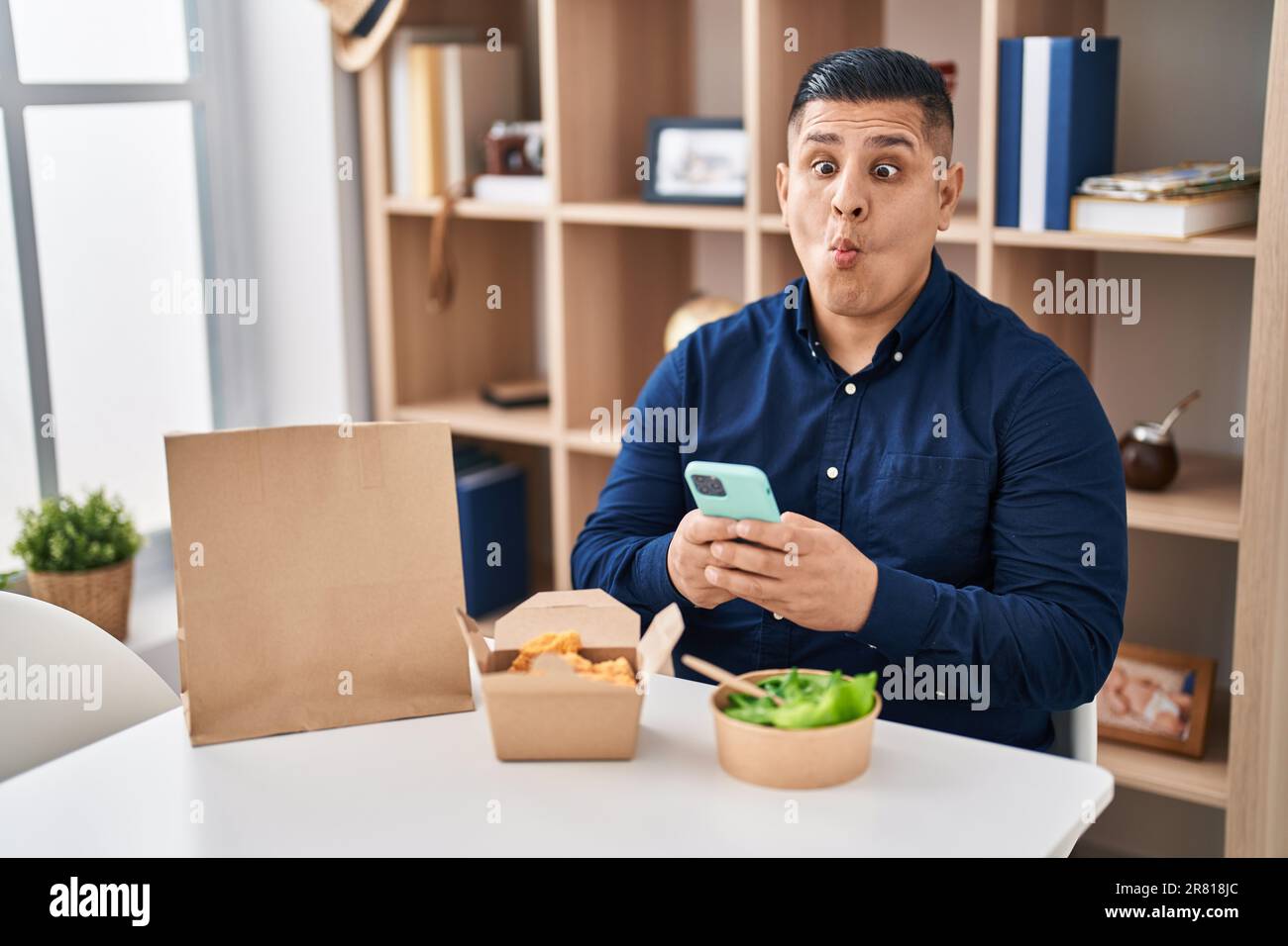 Hispanic young man eating take away food using smartphone making fish ...