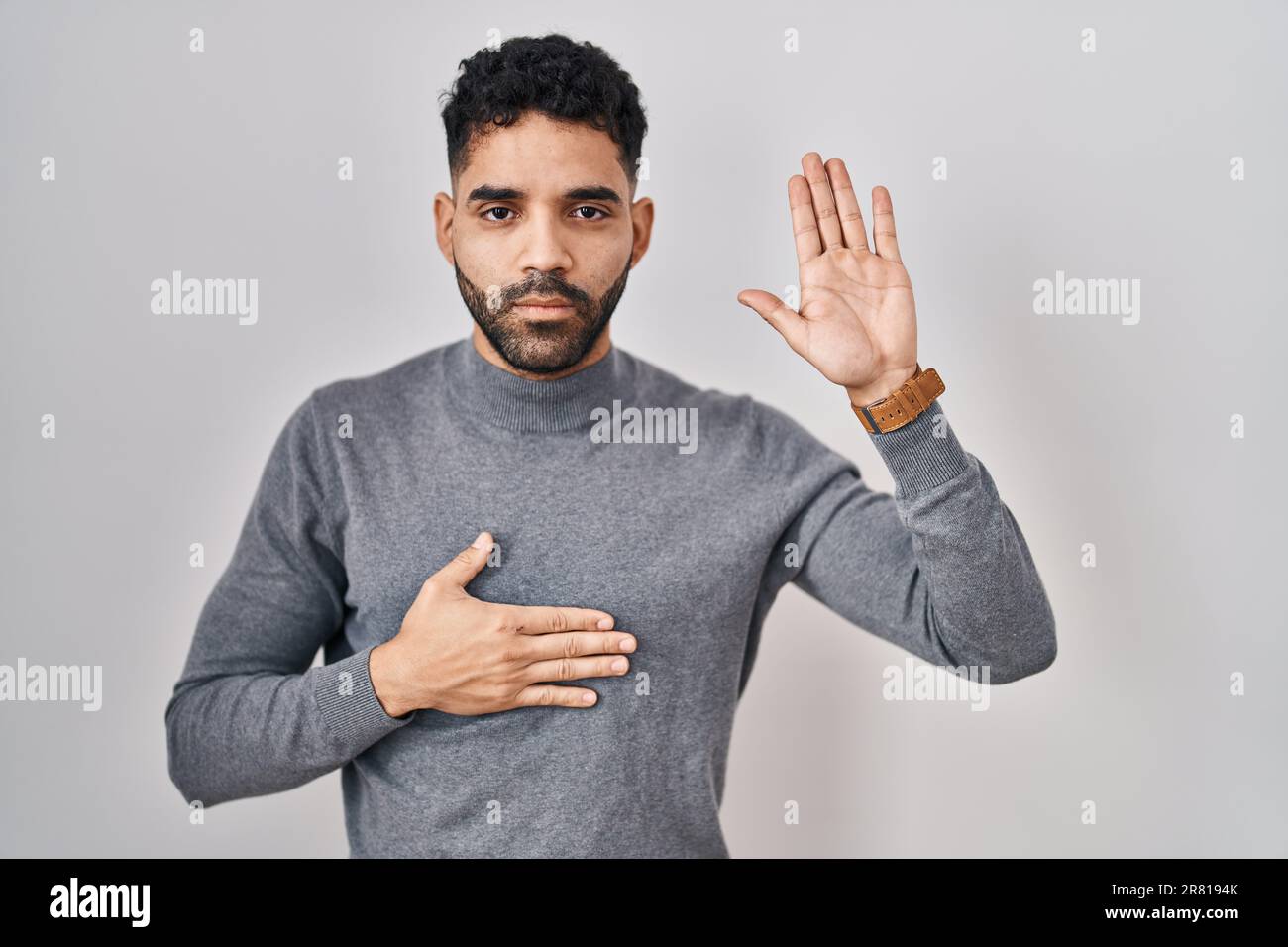 Hispanic man with beard standing over white background swearing with ...