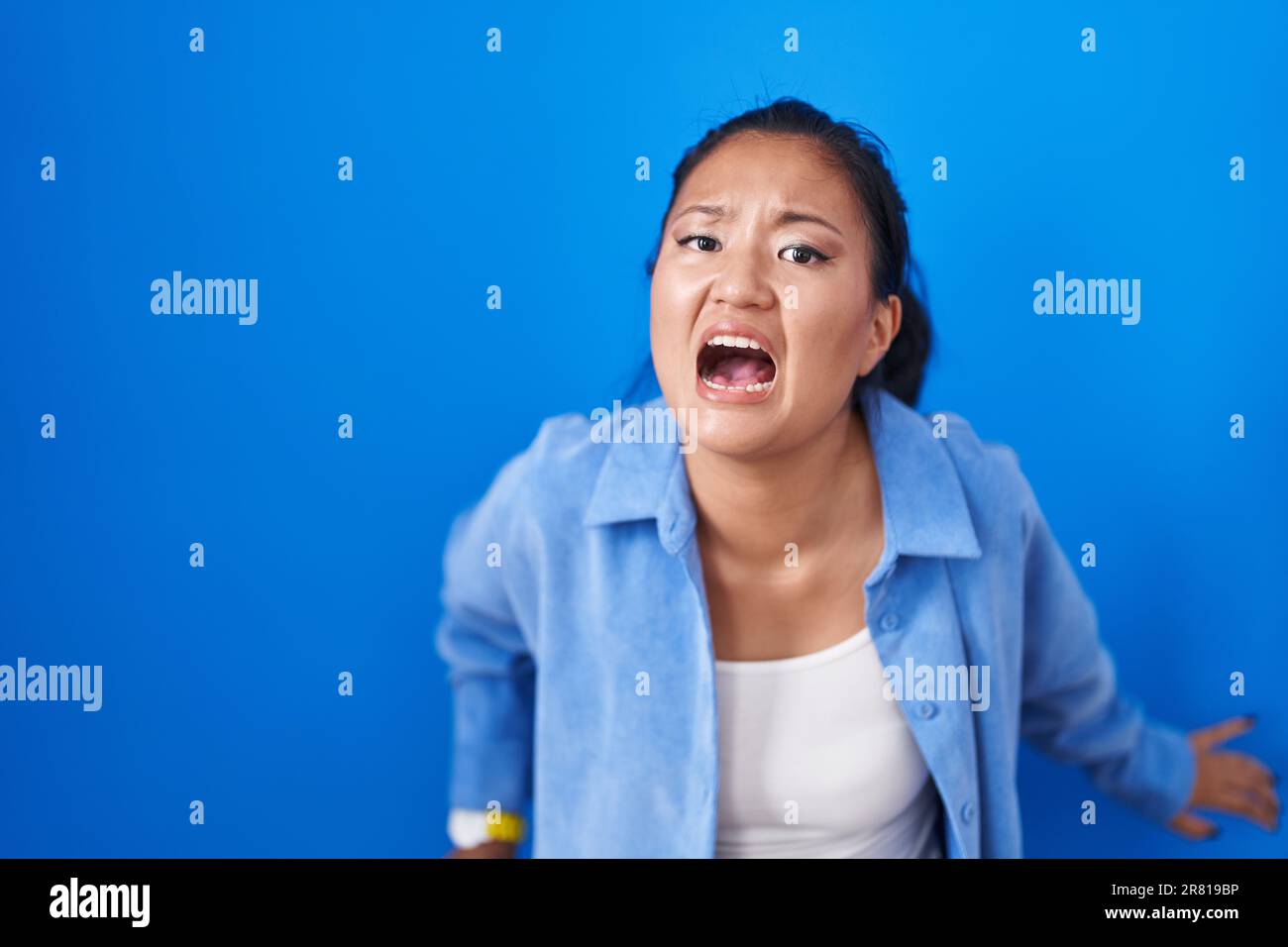 Asian young woman standing over blue background angry and mad screaming ...