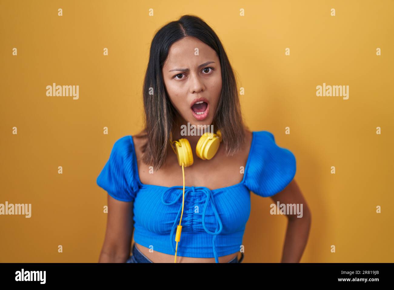 Hispanic young woman standing over yellow background in shock face ...