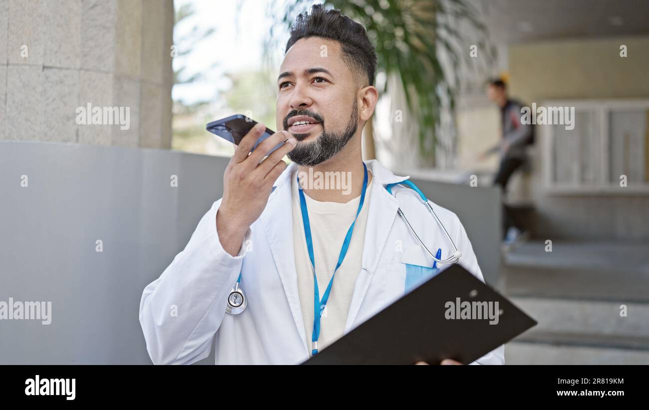 Young latin man doctor sending voice message by smartphone holding ...