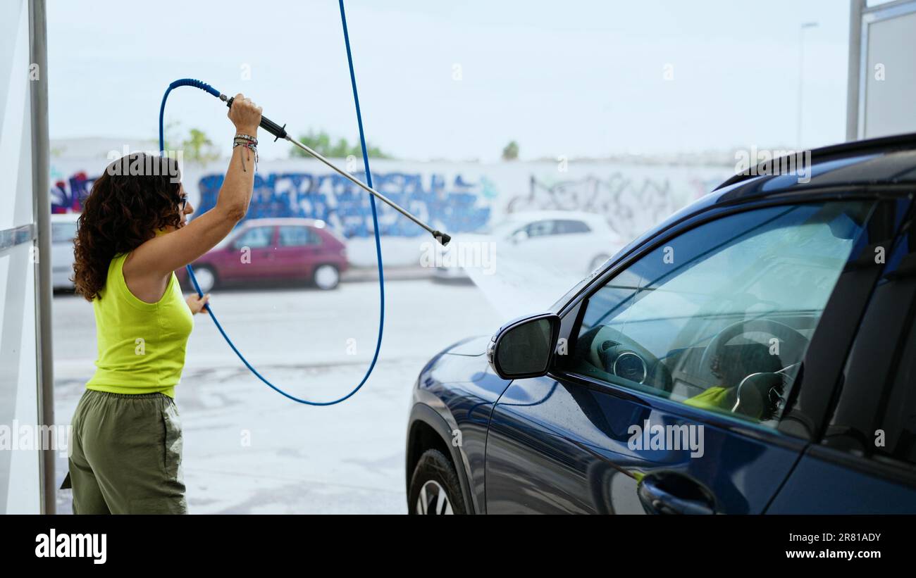 Middle age hispanic woman washing car with pressure washer at car wash ...