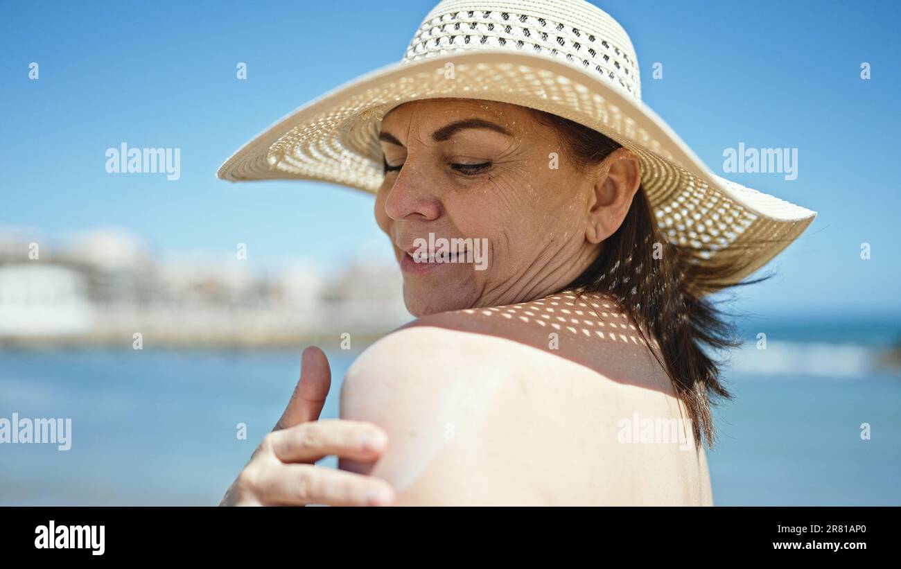 Middle age hispanic woman tourist smiling wearing summer hat applying ...