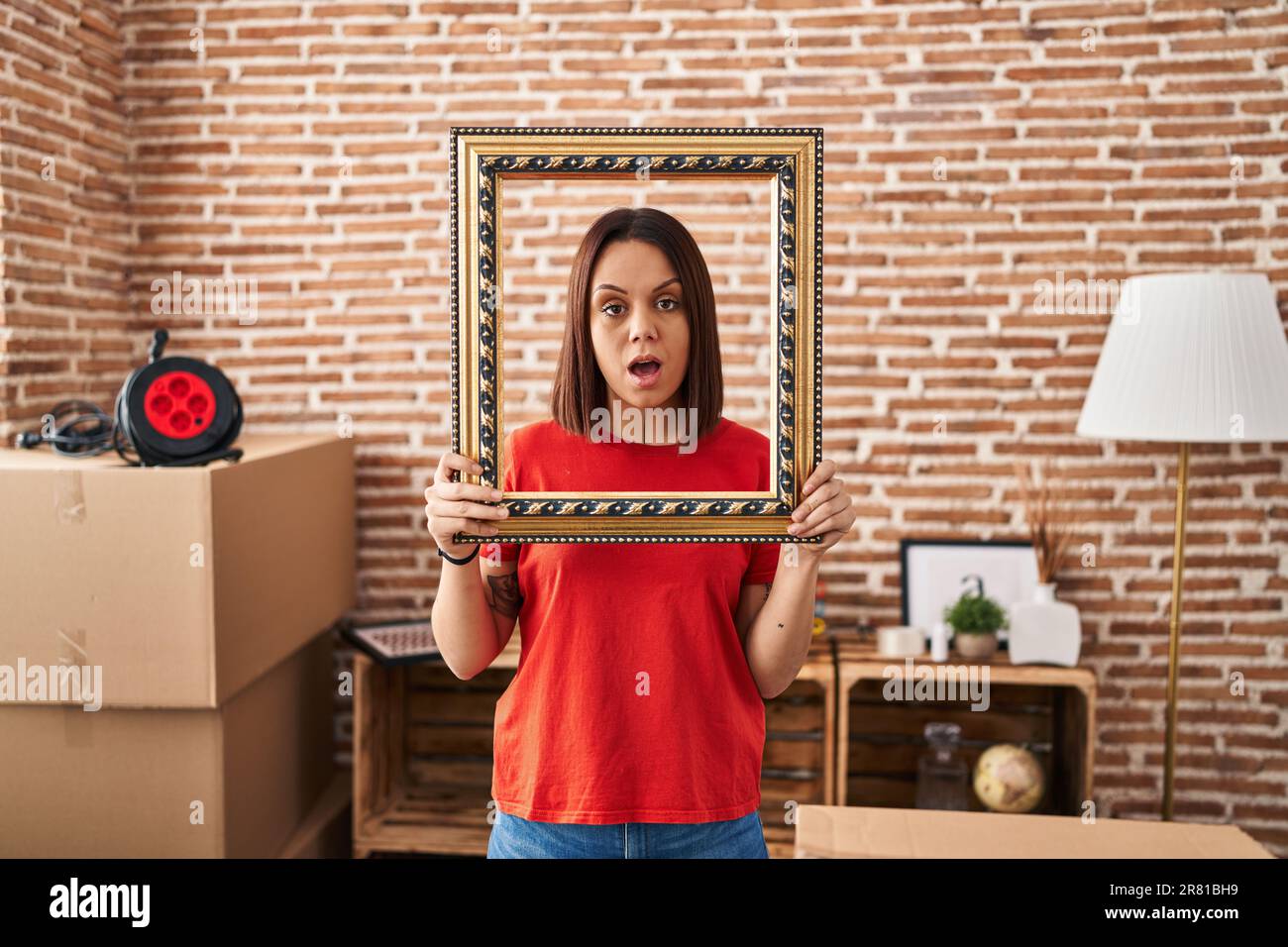 Young hispanic woman at new home holding empty frame in shock face ...