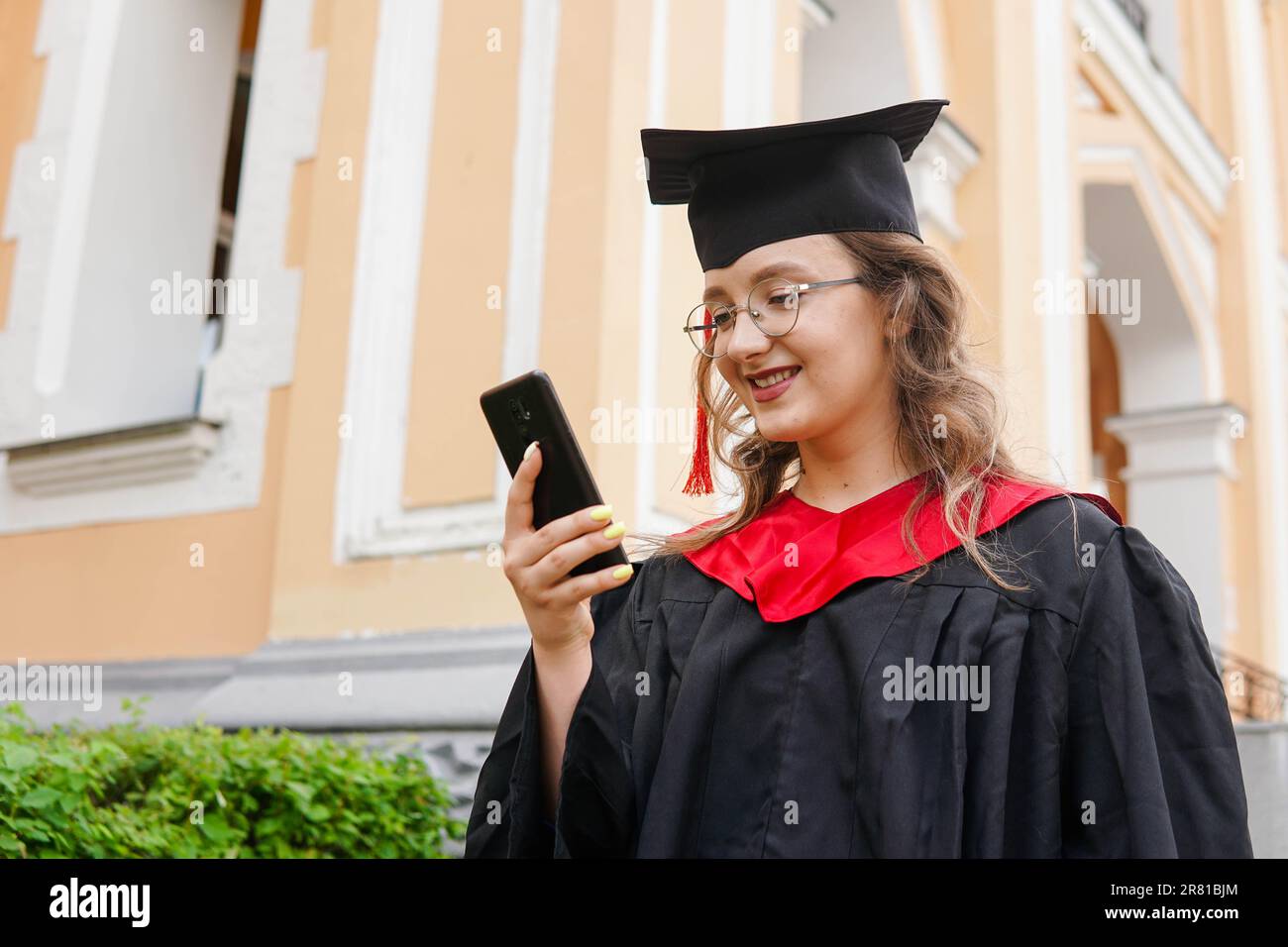 Happy smiling female graduate student in academic gown using smartphone ...