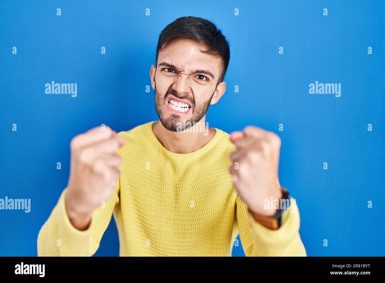 Hispanic man standing over blue background angry and mad raising fists ...