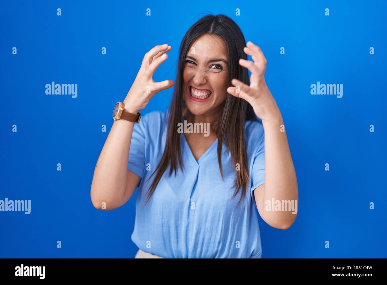 Young brunette woman standing over blue background shouting frustrated ...