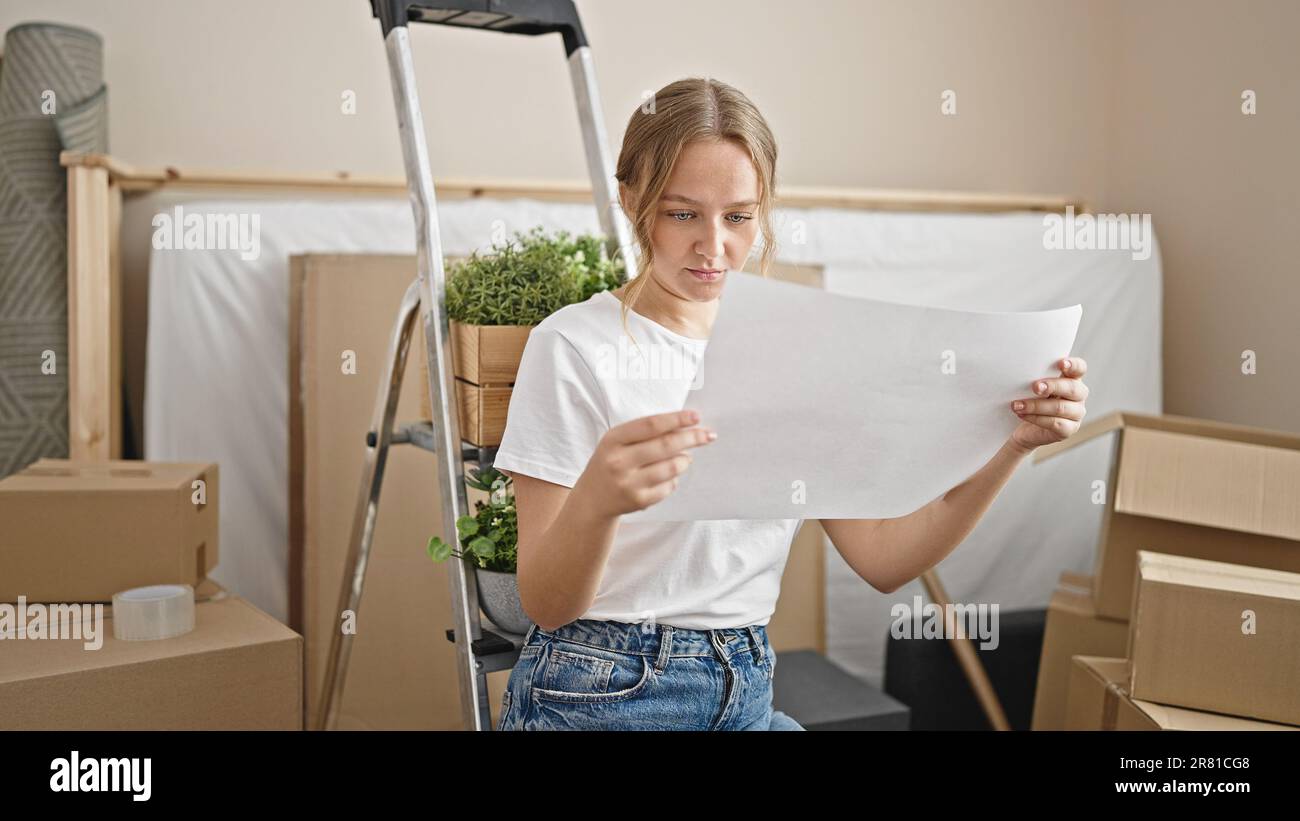 Young blonde woman sitting on ladder reading house project at new home ...