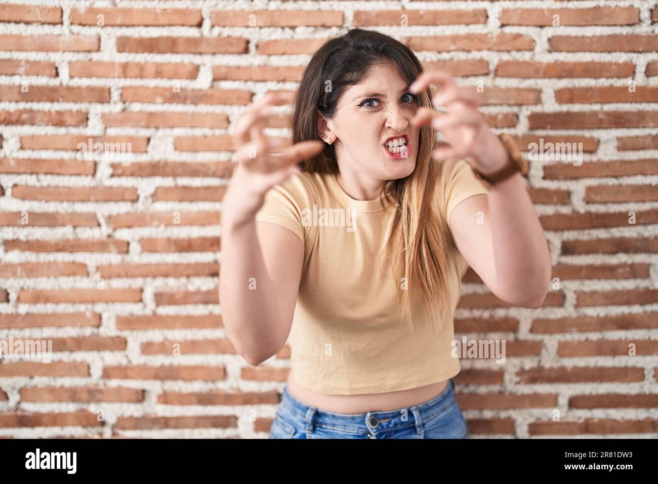 Young brunette woman standing over bricks wall shouting frustrated with ...