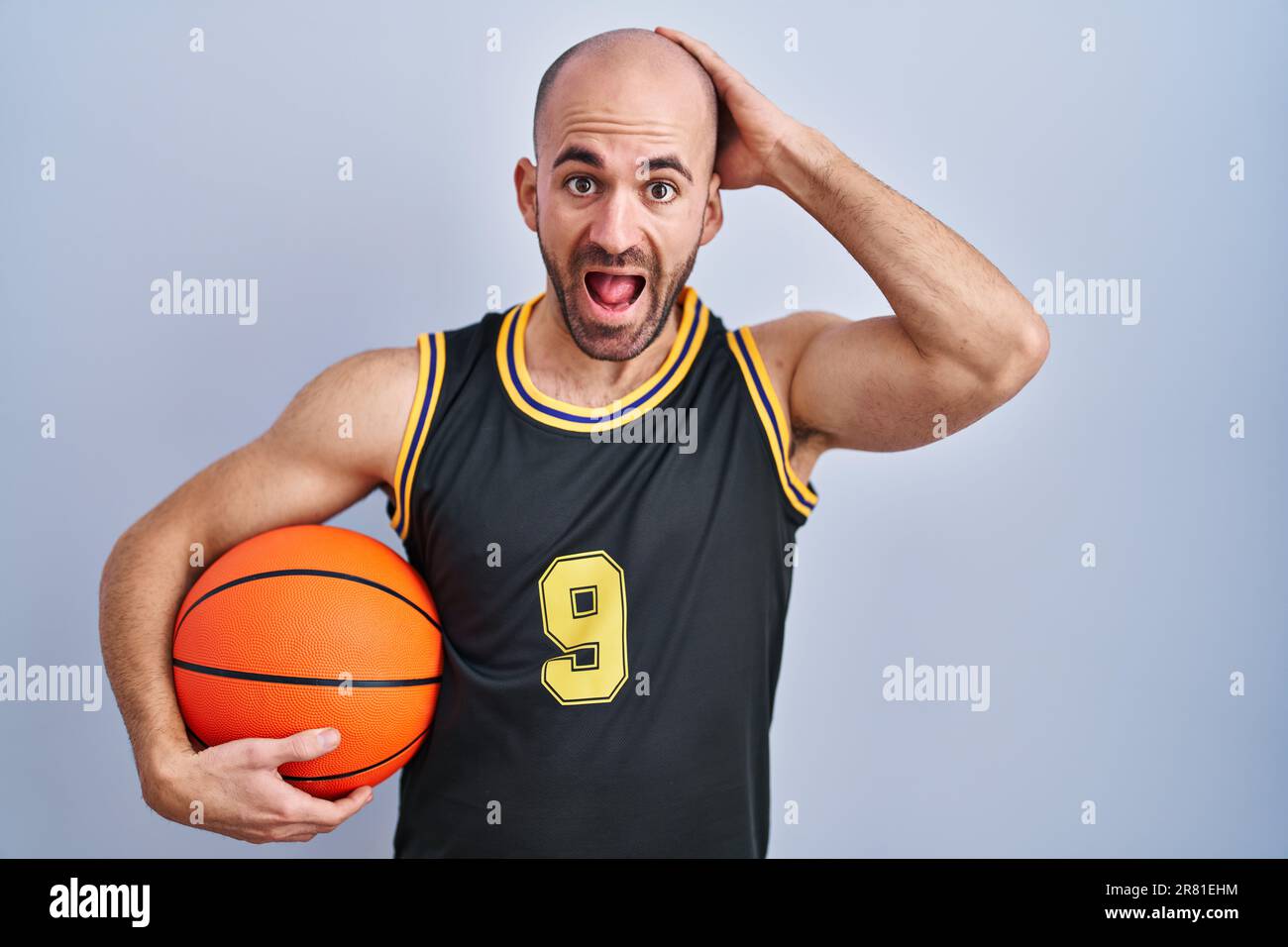 Young bald man with beard wearing basketball uniform holding ball crazy and scared with hands on ...