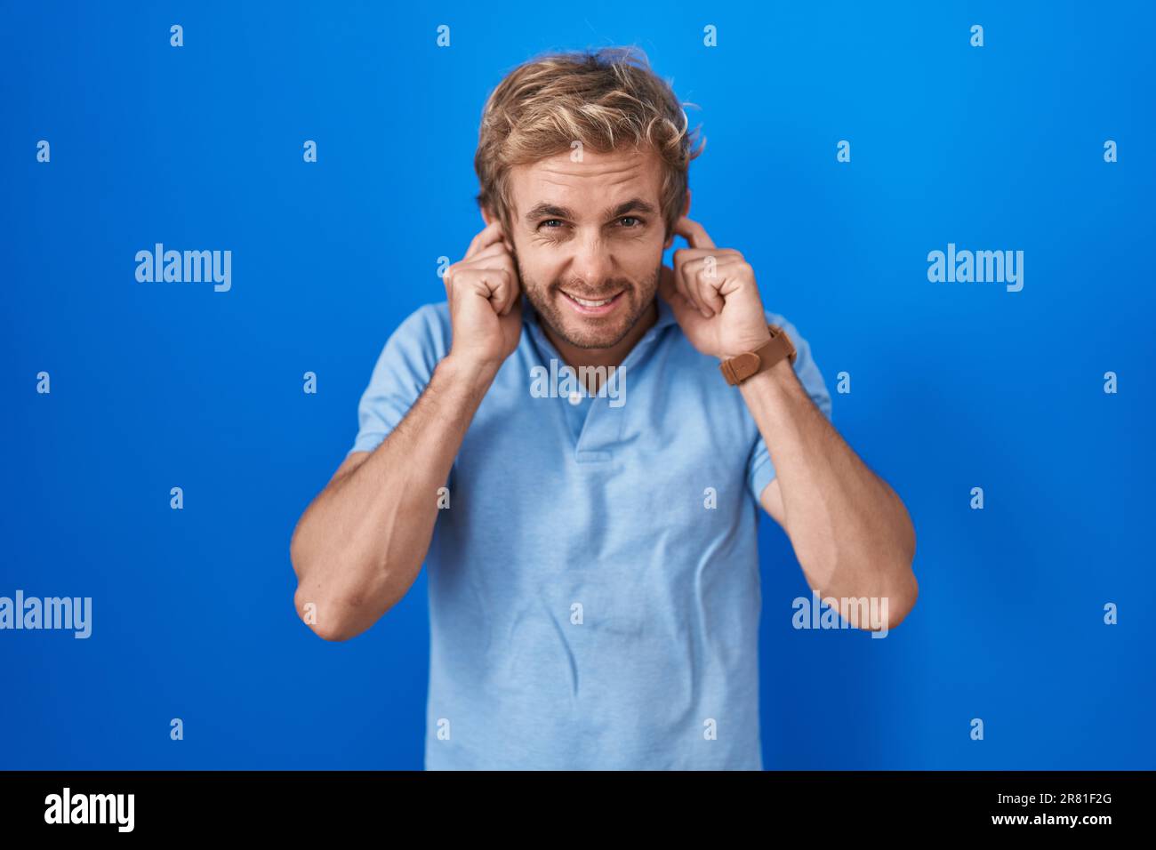 Caucasian man standing over blue background covering ears with fingers ...