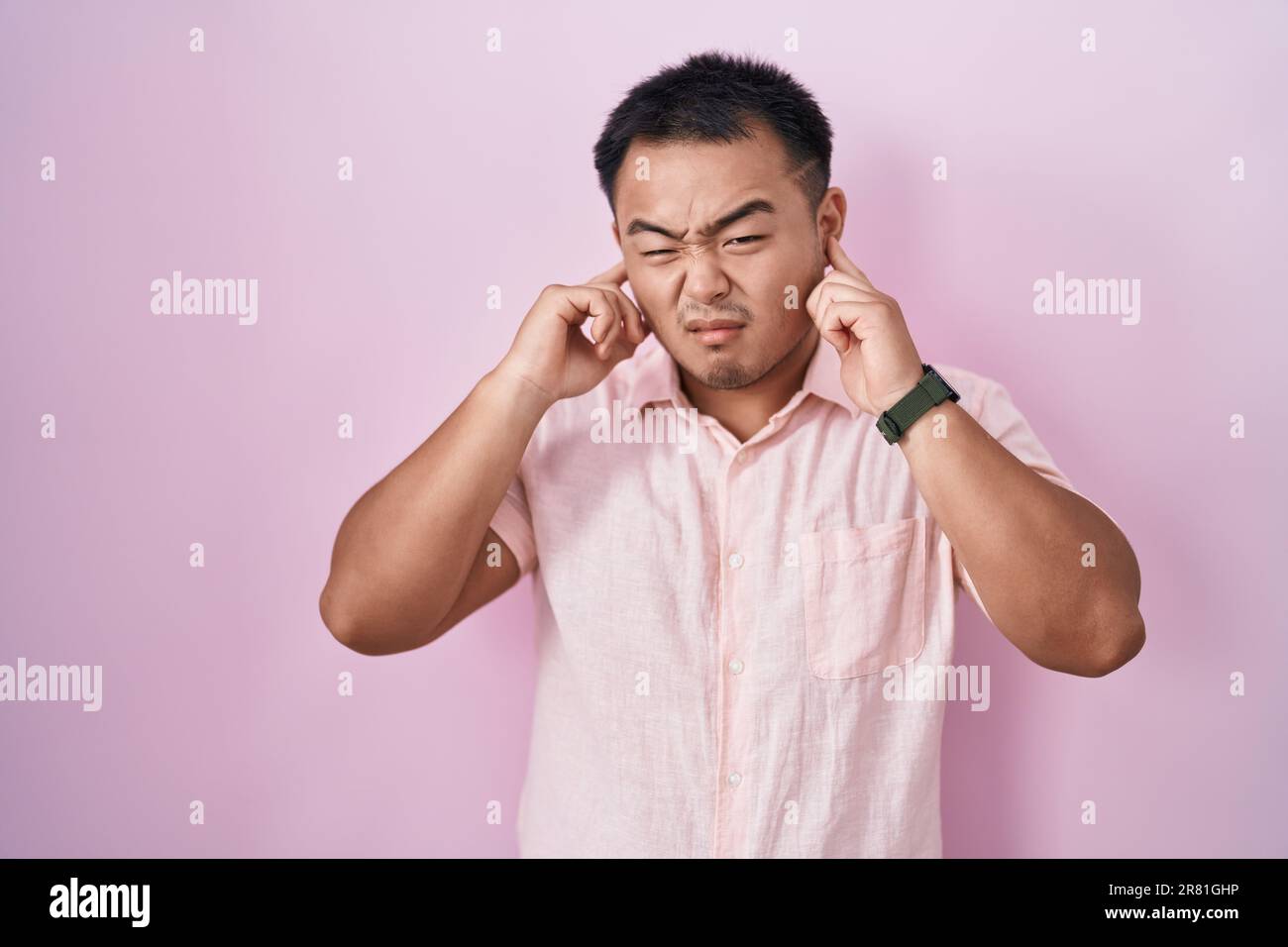 Chinese young man standing over pink background covering ears with ...