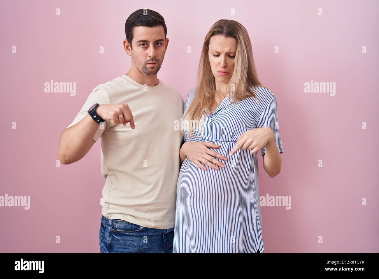 Young couple expecting a baby standing over pink background pointing ...