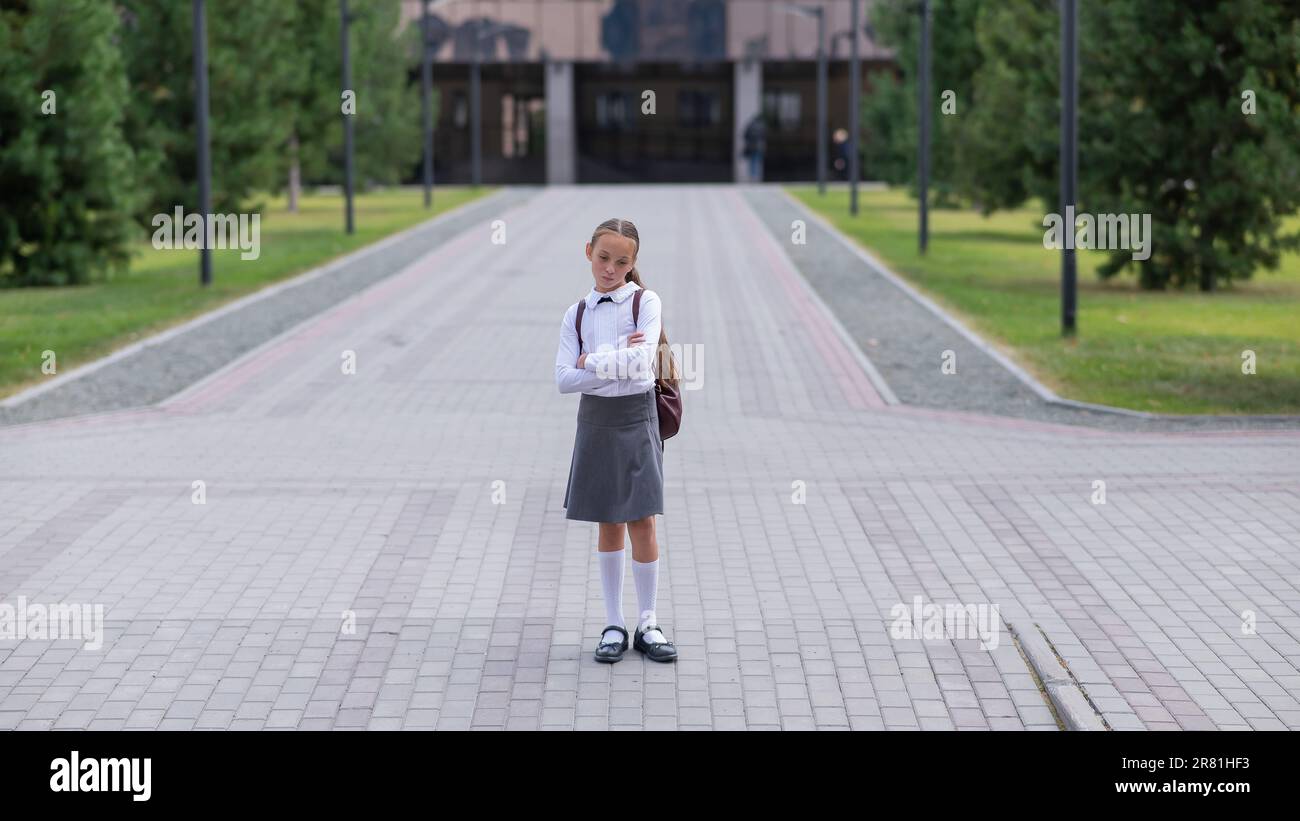 A sad caucasian girl in uniform and with a backpack holds her arms ...