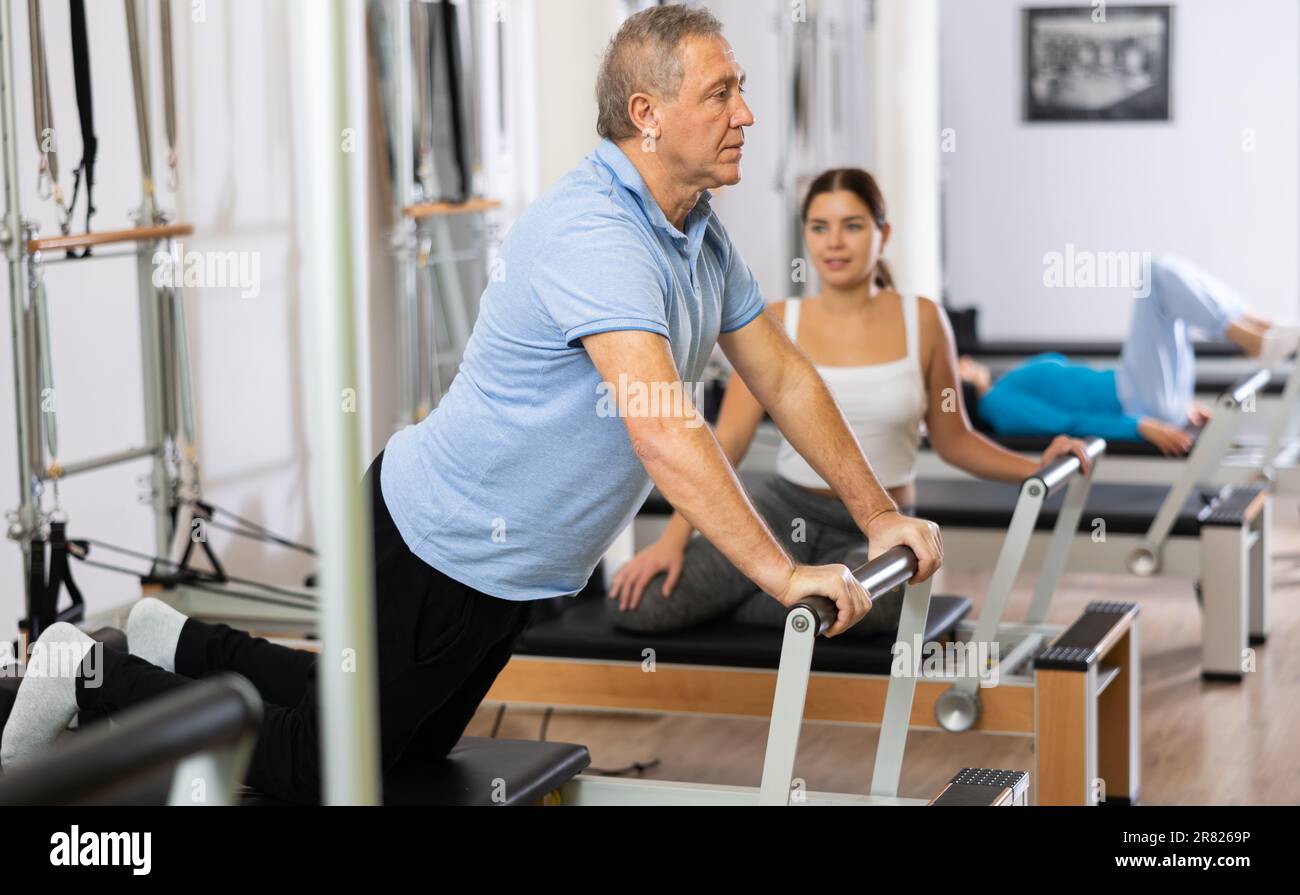 Focused old man performing stretching Pilates exercises with reformer ...