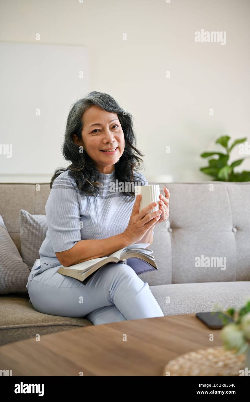 A portrait of a happy retired Asian woman in casual clothes sips coffee ...
