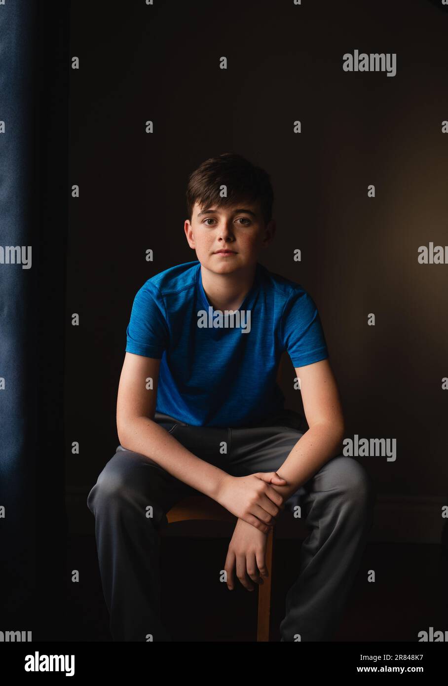 Portrait of tween boy with serious expression sitting in dark room ...