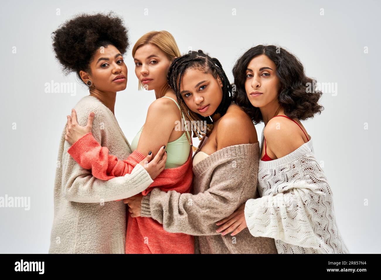 Portrait of confident and multiethnic women in knitted sweaters hugging ...